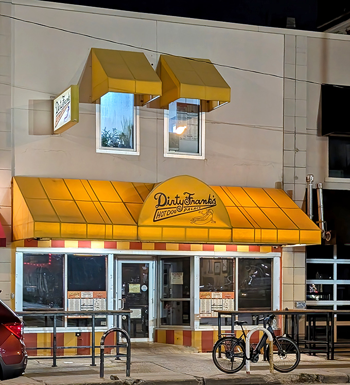 Night transforms Dirty Frank's into a golden hot dog temple, its yellow awnings glowing like the perfect mustard drizzle on a midnight snack.