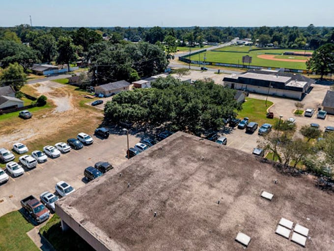 Overhead sun casts sharp shadows on a busy parking lot and the flat rooftop, overlooking a leafy suburban landscape that includes a distant baseball field.