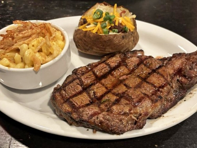 A perfectly grilled ribeye with textbook crosshatch marks, flanked by creamy mac and cheese and a loaded baked potato. The holy trinity of steakhouse indulgence.