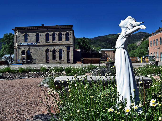 A stone bank building meets wildflower gardens where a white statue reaches skyward &ndash; history and beauty sharing the same mountain air.