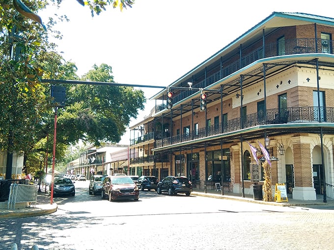 Bright morning sunlight illuminates the French Quarter-style architecture, featuring ornate iron balconies that line a busy, tree-shaded street corner under a clear blue sky.