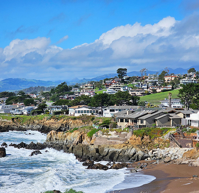 Sun-drenched houses perch on the dramatic California coast, with white-capped waves guarding the rocky shore below the bright, cloud-dotted sky.