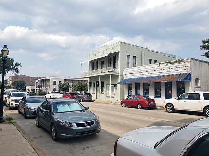 Soft daylight falls on a historic streetscape lined with pastel-colored buildings, including one with a classic iron balcony, as cars are parked along the curb under a lightly clouded sky.