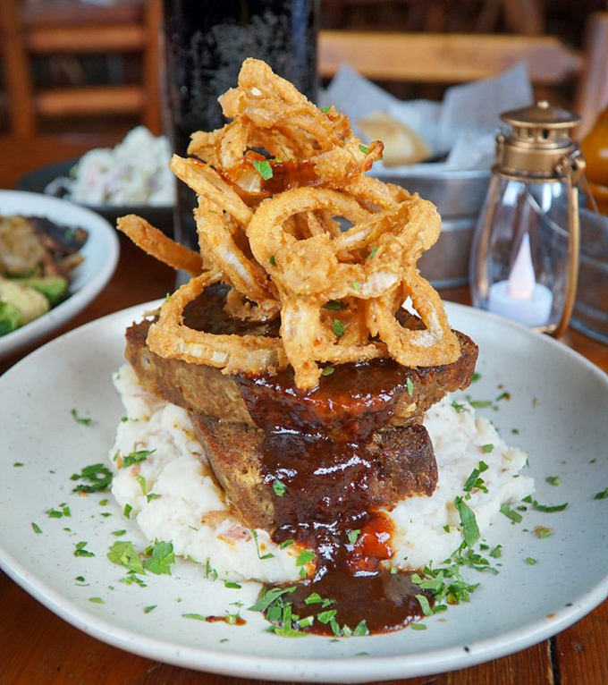 Elk meatloaf stacked like a delicious Jenga tower of wilderness flavor, crowned with crispy onion rings that defy gravity and restraint.