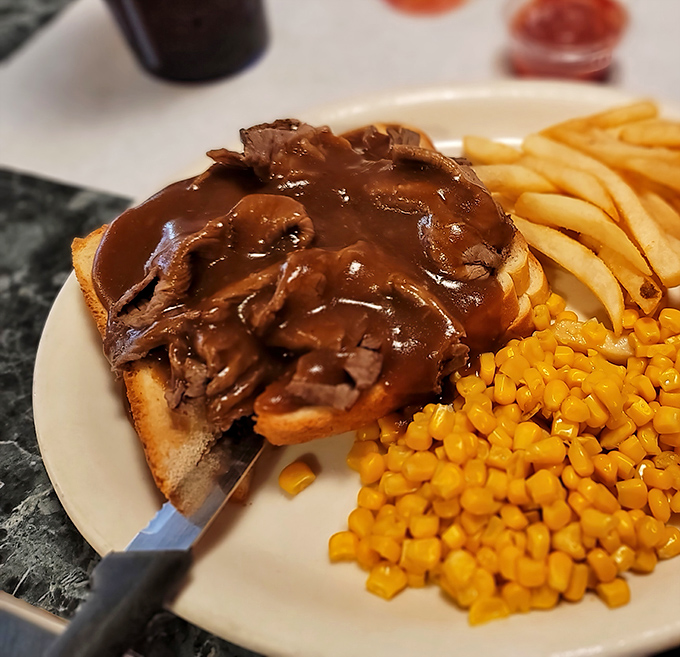 Hot beef sandwich with gravy that doesn't just sit on the plate &ndash; it commands respect, surrounded by its loyal subjects: fries and corn.