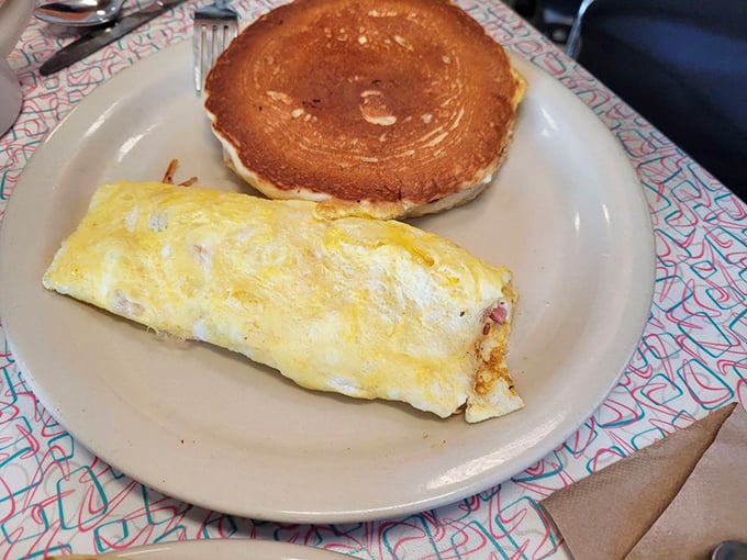 The breakfast power couple that never disappoints: a perfectly executed omelet alongside a golden pancake, served on those distinctive placemats that scream "classic diner."