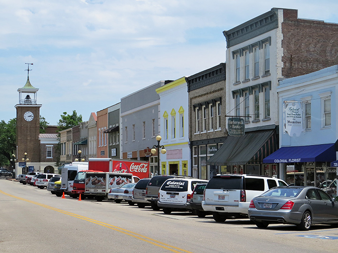 Bright daylight illuminates a line of historic, multi-colored storefronts along a busy downtown street, leading the eye toward the distinctive clock tower under a partially cloudy sky.
