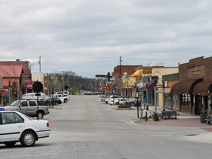 Overcast clouds hang above a classic small town main street, lined with low-slung brick buildings and parked cars, leading toward the far end of the quiet downtown.