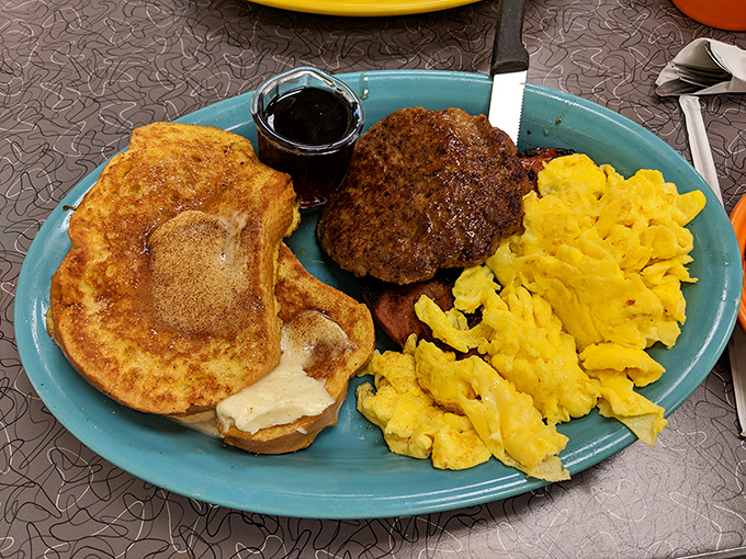 Breakfast indecision solved: golden French toast, scrambled eggs, and a perfectly charred sausage patty&mdash;the breakfast trinity on a single blue plate.