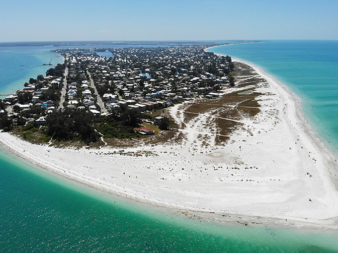 Blinding white sands and turquoise waters meet at the point of a barrier island community, where houses are nestled between the ocean and the bay under the bright, cloudless sun.