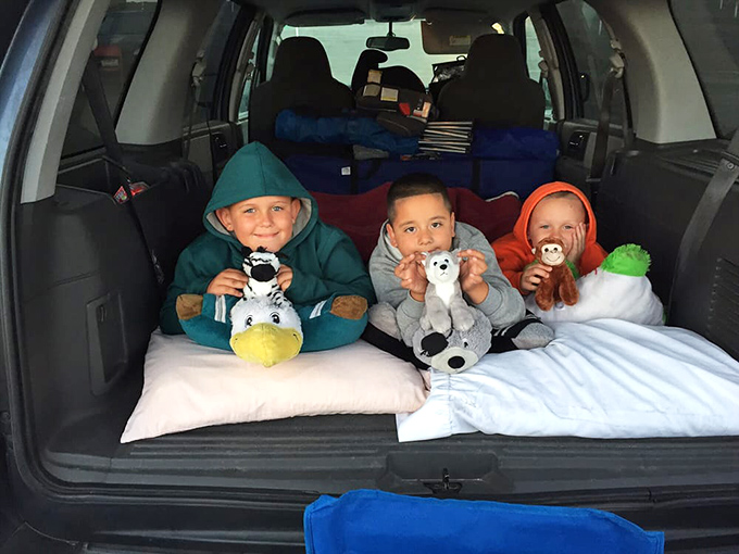 Three young movie enthusiasts demonstrate the ultimate drive-in comfort hack: pillows, stuffed animals, and the perfect blanket nest in the back of an SUV.