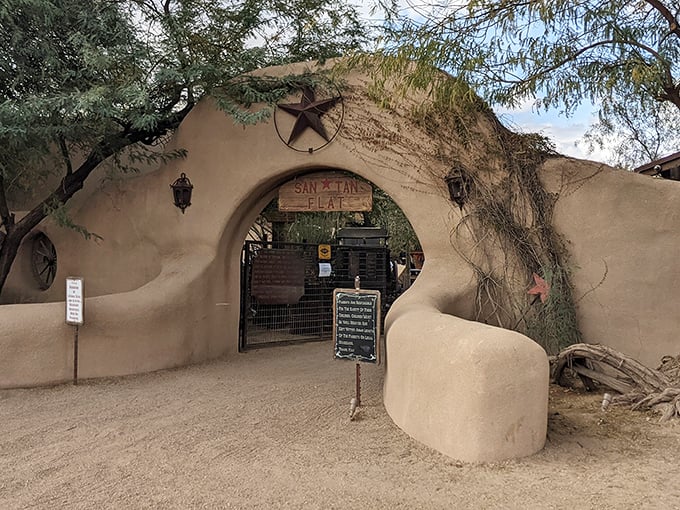An arched adobe gateway marked with a rustic wooden sign and metal star invites visitors into San Tan Flat's desert oasis atmosphere.