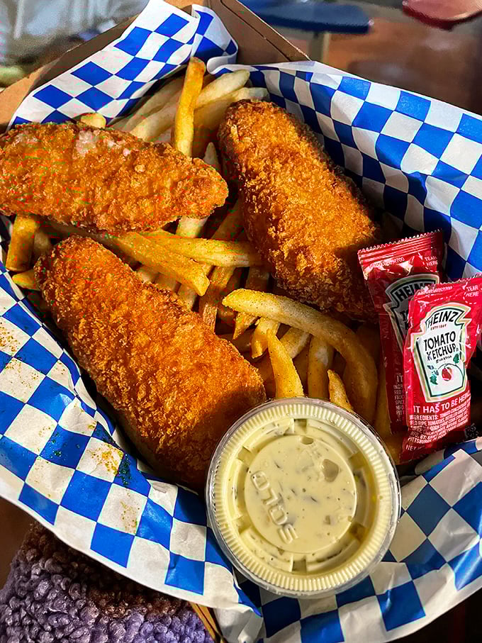 Golden-battered fish and chips arrive in their blue checkered paper boat—crispy vessels sailing through seas of tartar sauce and ketchup.