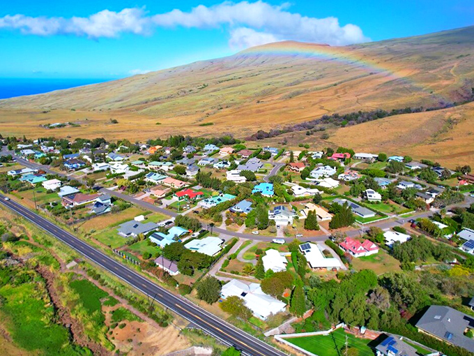 A sunlit Waimea community stretches across green foothills, framed by sweeping open plains and a soft rainbow arching under Hawaii&rsquo;s bright blue sky.