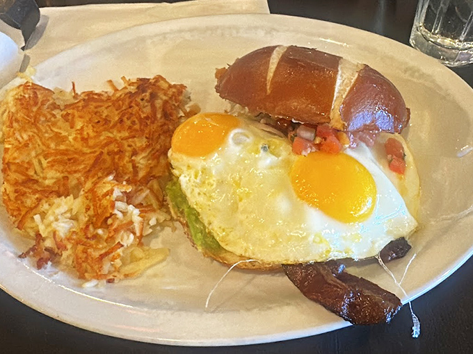Breakfast sandwich perfection: sunny-side-up eggs perched atop avocado on a pretzel bun with crispy hash browns standing guard. Morning has officially broken good.