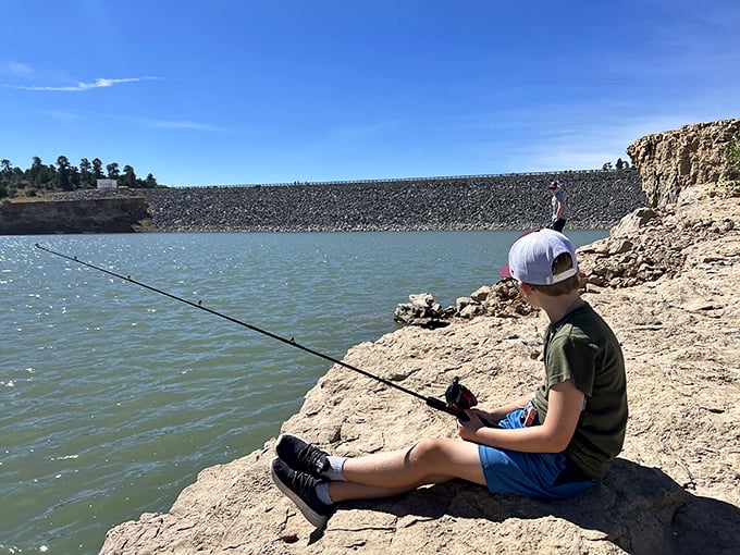 This young angler demonstrates the timeless art of patience, perched on nature's own recliner while the dam stands guard in the background.