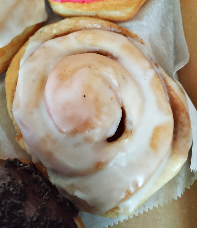 The cinnamon roll glistens with vanilla glaze, its perfect spiral promising the kind of comfort that makes Monday mornings bearable.