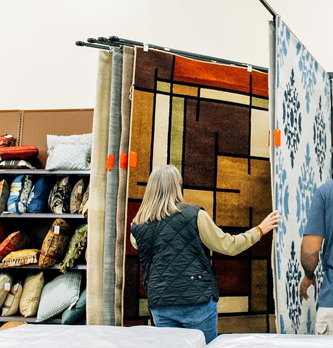 A customer contemplates carpet choices that could transform a room. The geometric patterns whisper of mid-century modern without the mid-century prices.