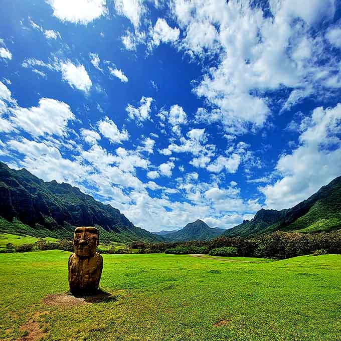 Under a canvas of scattered clouds, a carved figure stands sentinel in Kahana Valley, where ancient traditions meet breathtaking landscapes.