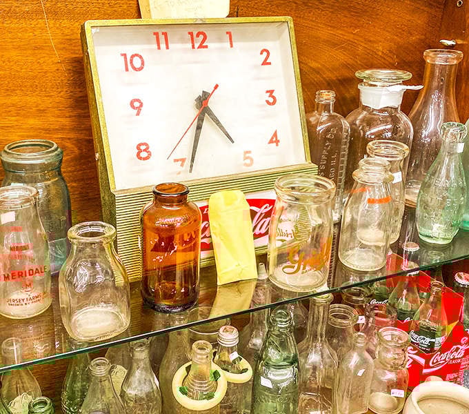 A vintage clock keeps time among glass bottles that once delivered milk to Anchorage doorsteps, each one a transparent vessel of local history.