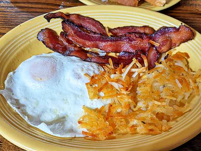 The holy trinity of breakfast perfection – crispy bacon, golden hash browns, and an egg cooked just right on a no-nonsense yellow plate.