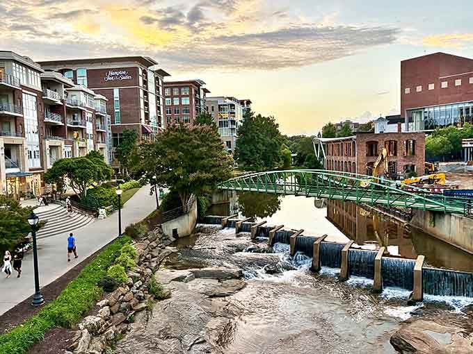 Sunset illuminates Greenville's Reedy River Falls with modern apartments and a green pedestrian bridge creating urban tranquility.