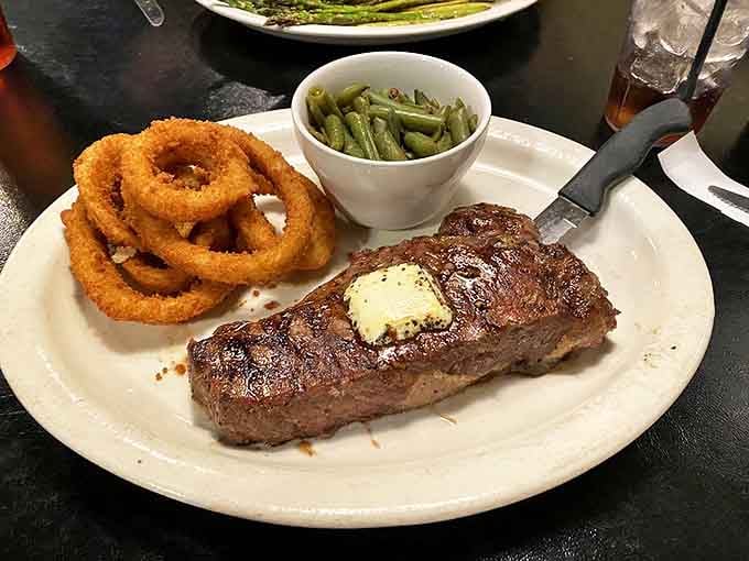 A perfectly seared KC Strip steak with a pat of melting butter, accompanied by golden onion rings and green beans &ndash; the holy trinity of steakhouse perfection.