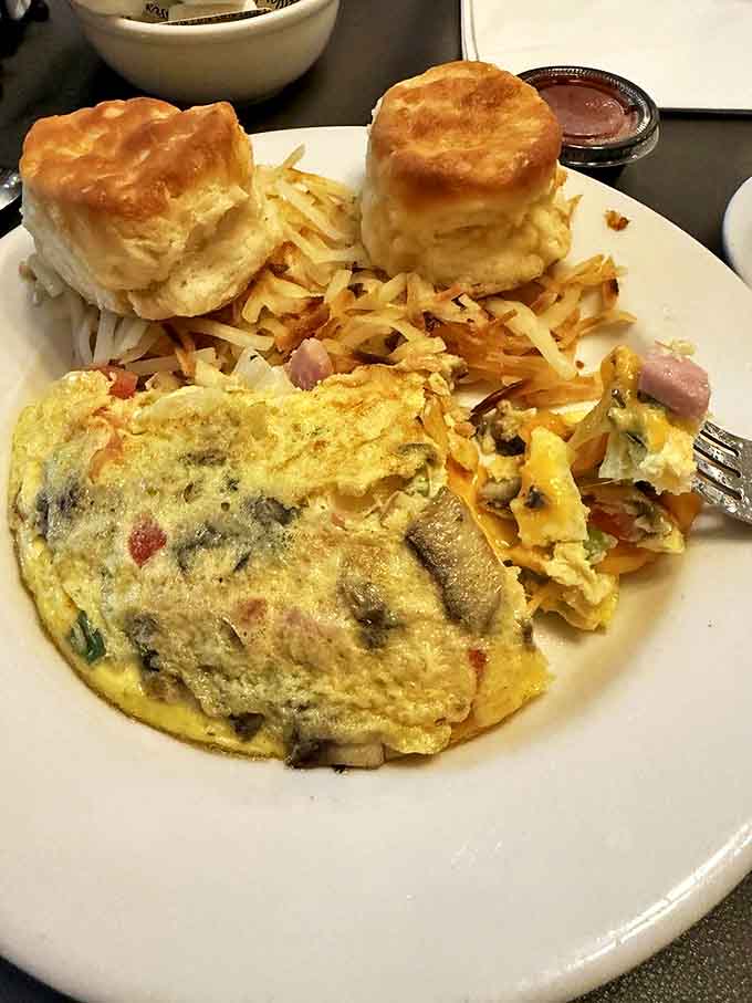 The omelet arrives like a fluffy yellow pillow stuffed with treasures, flanked by biscuits so tall they practically need their own zip code.