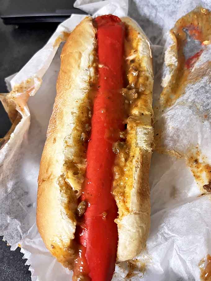 The hot dog arrives like a splash of color against the white wrapper, proof that sometimes the simplest pleasures deliver the most profound satisfaction.