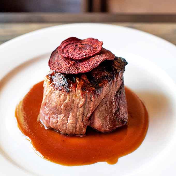 This filet mignon sits in its sauce like royalty on a throne, topped with what appears to be beet chips—a crown for beef nobility.