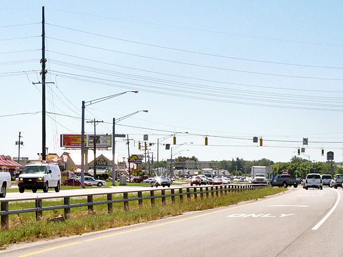 A bustling day in Kokomo shows cars traveling along the sunny highway, passing local businesses and bright billboards under clear skies.