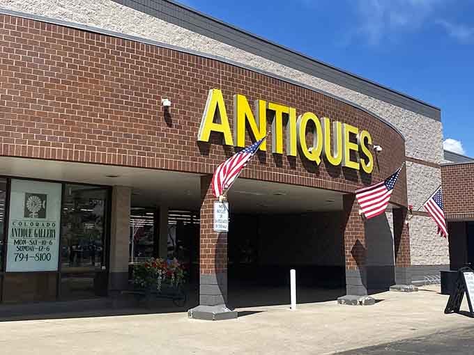 The welcoming entrance of Colorado Antique Gallery beckons with bright yellow signage and patriotic flags, promising treasures inside for curious shoppers.