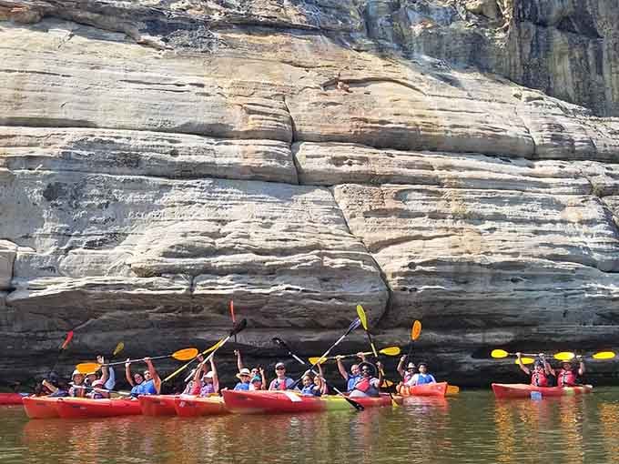 Paddlers in bright red kayaks explore towering sandstone cliffs that rise dramatically from the river's edge below.