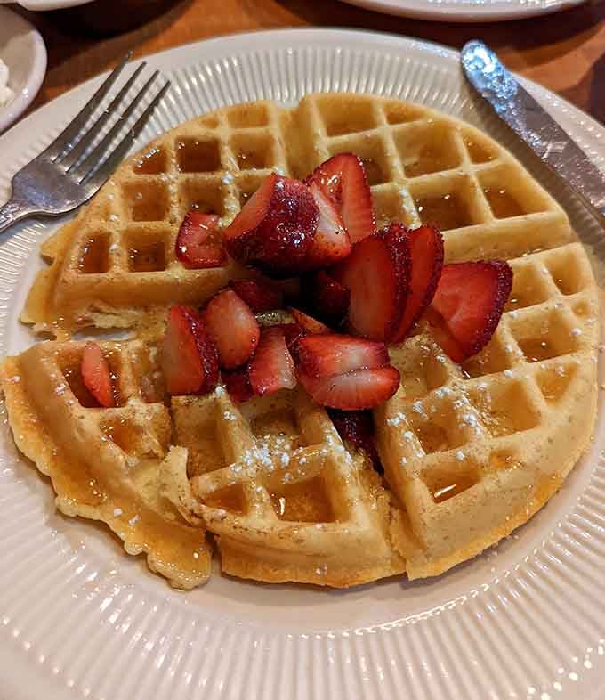 Belgian waffle crowned with strawberries like breakfast royalty, those deep pockets perfectly engineered to hold maximum syrup without structural failure.