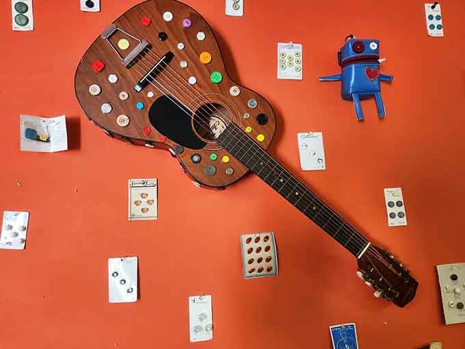 An acoustic guitar casually decorated with colorful buttons hangs on the orange wall like it's perfectly normal.