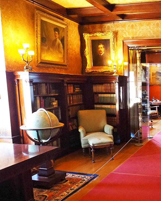 The library features floor-to-ceiling bookshelves and family portraits, creating an atmosphere of intellectual refinement and old money.