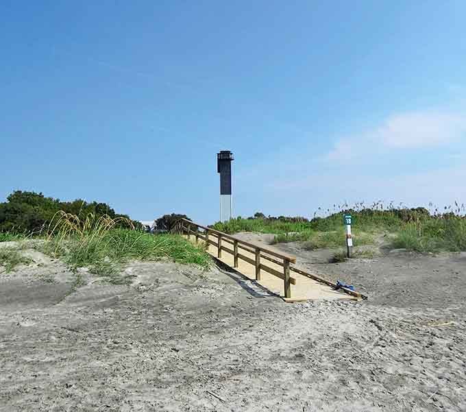 The beach access path leads toward endless sand and surf, with the lighthouse standing guard like a very tall, very angular lifeguard on duty.