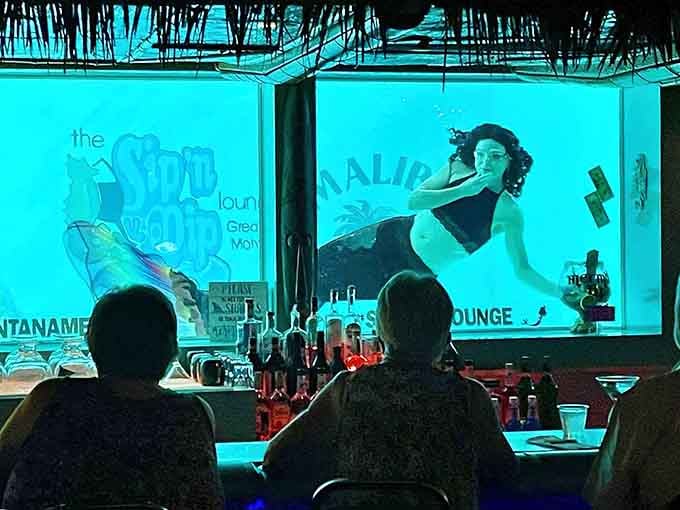 Behind the bar's blue-lit windows, mermaids glide through water while patrons sip cocktails in tiki paradise.