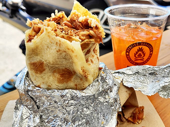 Wrapped like a present and opened like treasure, this foil-bundled burrito reveals its golden-tortilla glory alongside a spicy orange beverage companion.