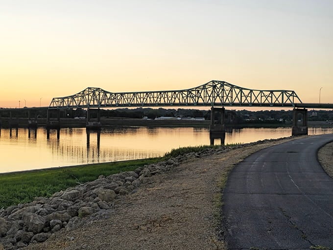 Golden sunset light reflects off the quiet river as you stroll along the paved path toward this grand truss bridge.