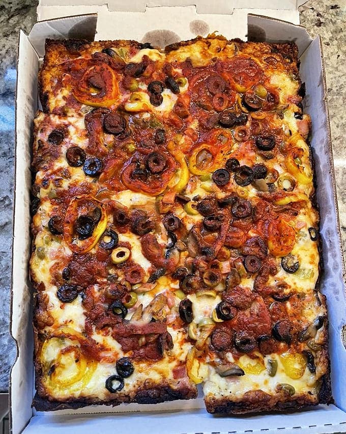 Detroit-style pizza in a rectangular pan, loaded with toppings that create a landscape of melted cheese perfection.