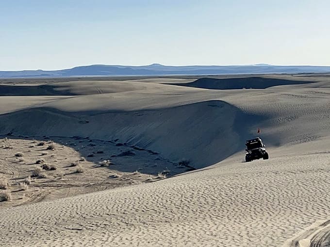 An off-road vehicle perches atop a massive dune overlooking the vast sandy expanse and distant mountains beyond.