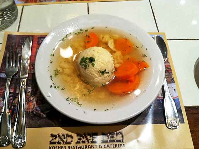 The matzo ball soup arrives like liquid sunshine, with that perfect dumpling floating majestically in broth clear enough to read your fortune.