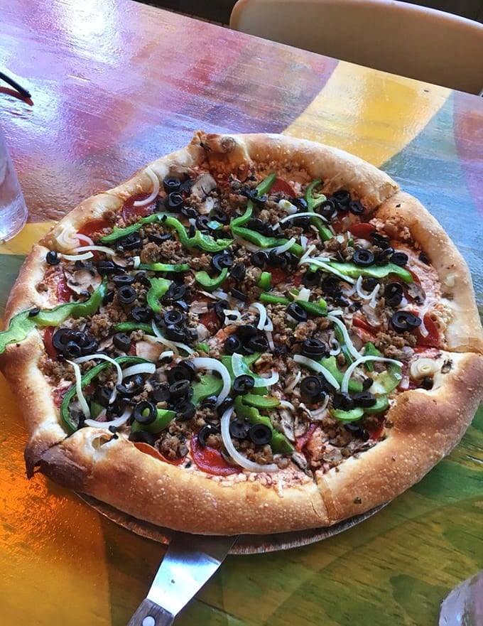 A vegan pizza loaded with peppers, olives, and possibilities, served on a table that's basically a rainbow threw a party.