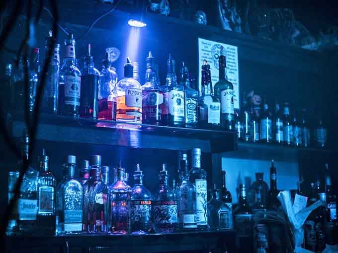 Behind that bar, bottles gleam under blue light like potions waiting to cast their delicious spells on you.