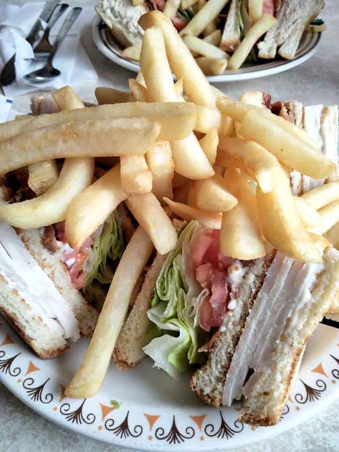 The club sandwich that requires jaw exercises before attempting. Layers of turkey, crisp lettuce, and tomato create the skyscraper of sandwiches, with fries as the perfect foundation.