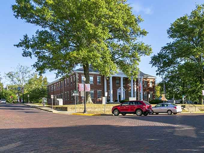 Mature trees shade the courthouse approach, their canopy creating a natural gateway to this stately brick building with white columns.