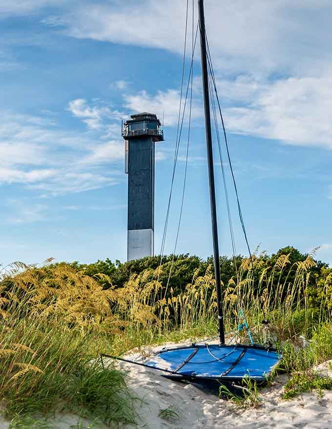 A beached sailboat rests among sea oats, the lighthouse rising behind like a patient friend waiting for the next high tide adventure.