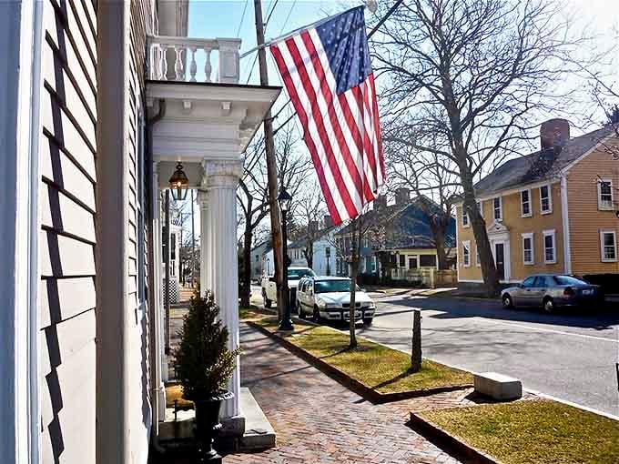 A patriotic American flag waves proudly over Wickford Village's historic main street, where colonial buildings create perfect small-town atmosphere.