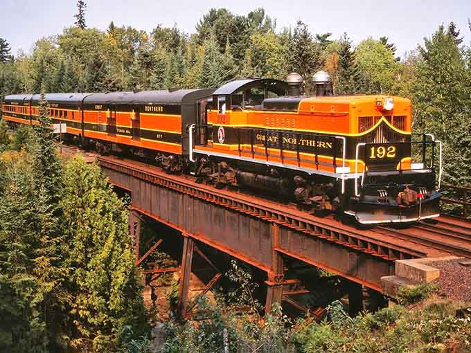 That striking orange and black locomotive crossing the trestle looks like it stepped straight out of a vintage postcard.
